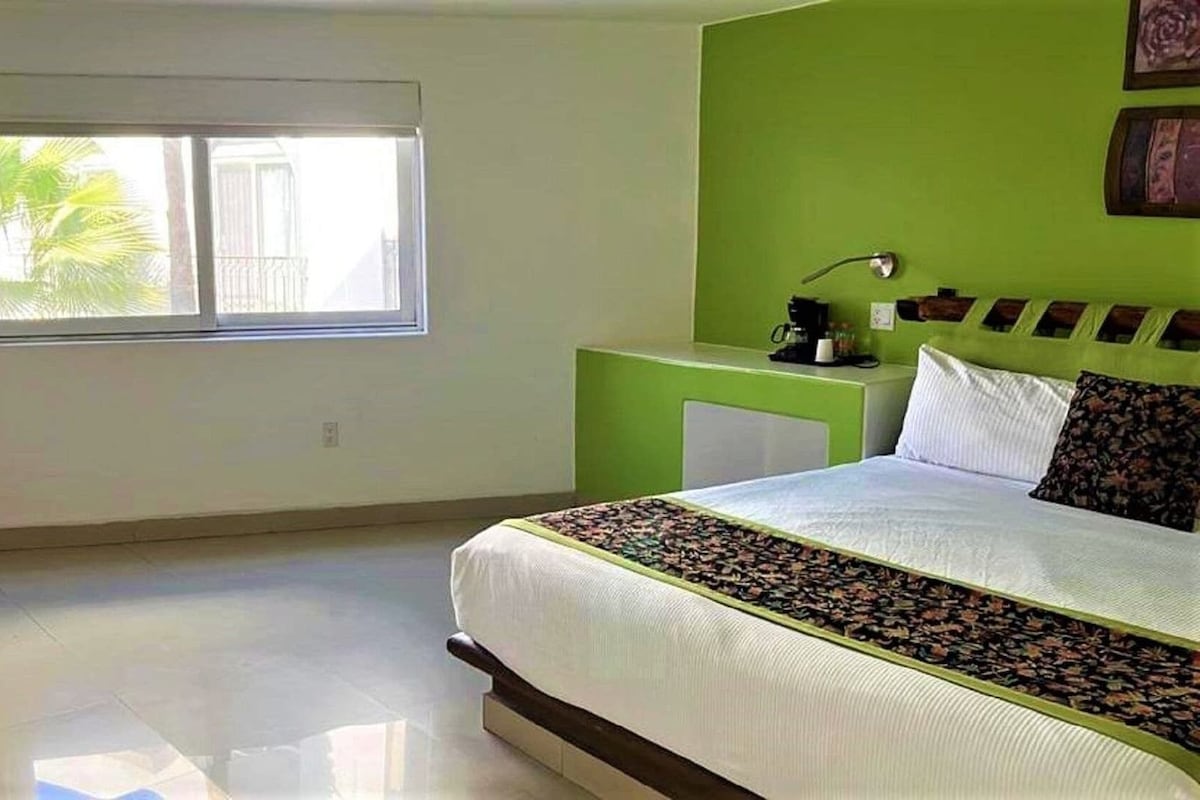 Quarto com cama, janela, mesa de cabeceira e luminária, e uma parede verde como detalhe.