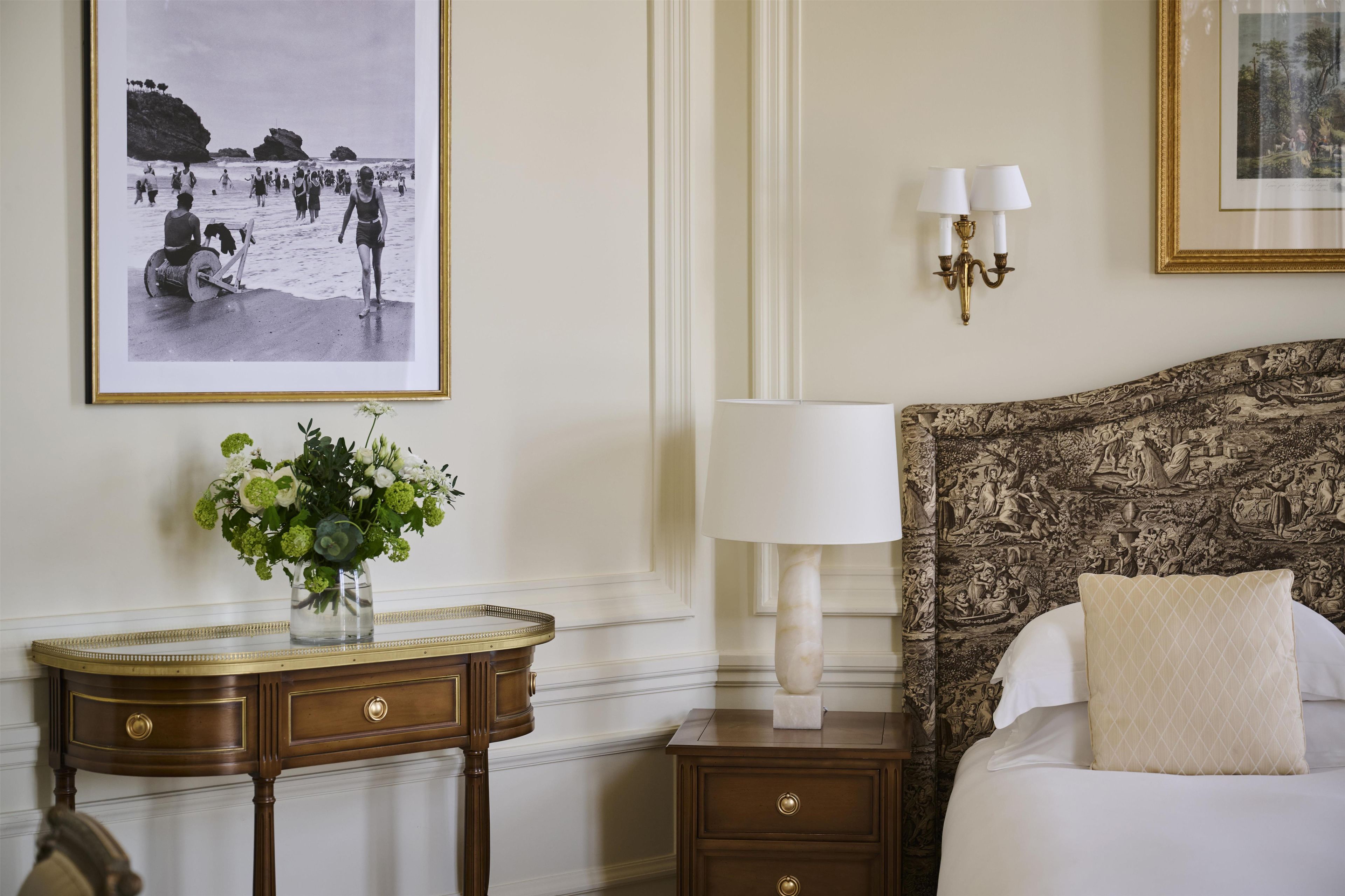 Quarto de hotel com uma cama, uma mesa de cabeceira com abajur, uma fotografia emoldurada na parede e um vaso com flores na mesa de cabeceira.