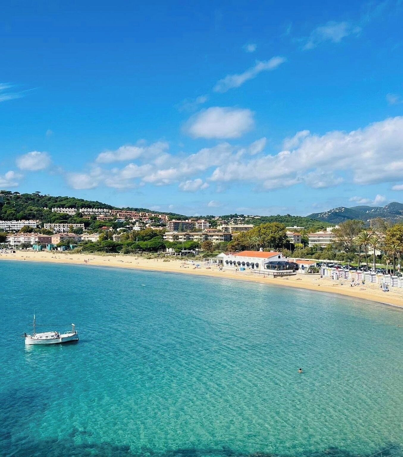 Uma cidade costeira com mar azul cristalino, uma praia arenosa e edifícios ao fundo.