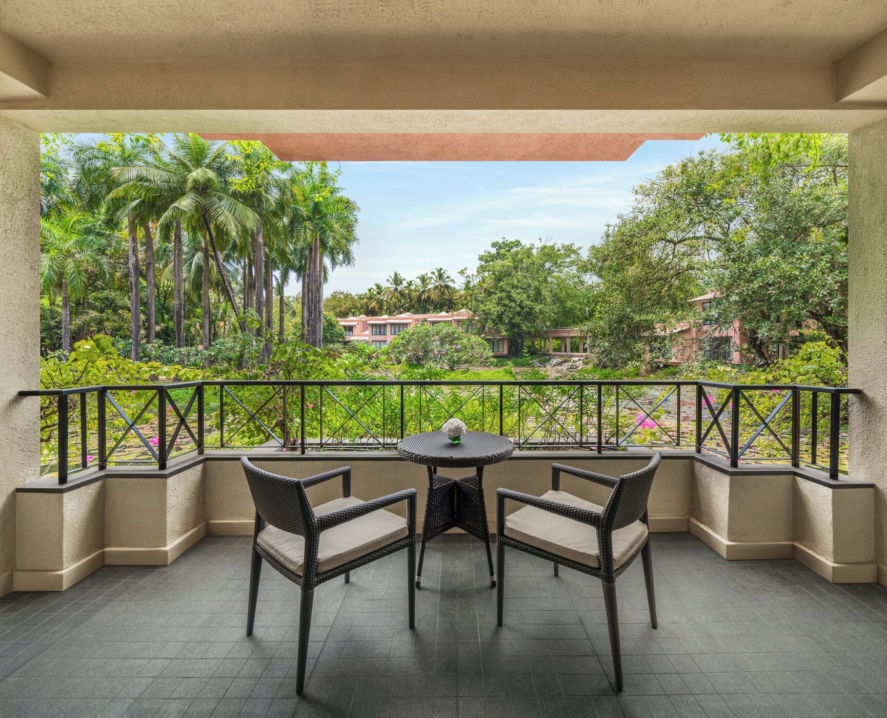 Uma área de estar coberta ao ar livre, com mesa e cadeiras, cercada por um corrimão, com vista para um jardim exuberante e edifícios.
