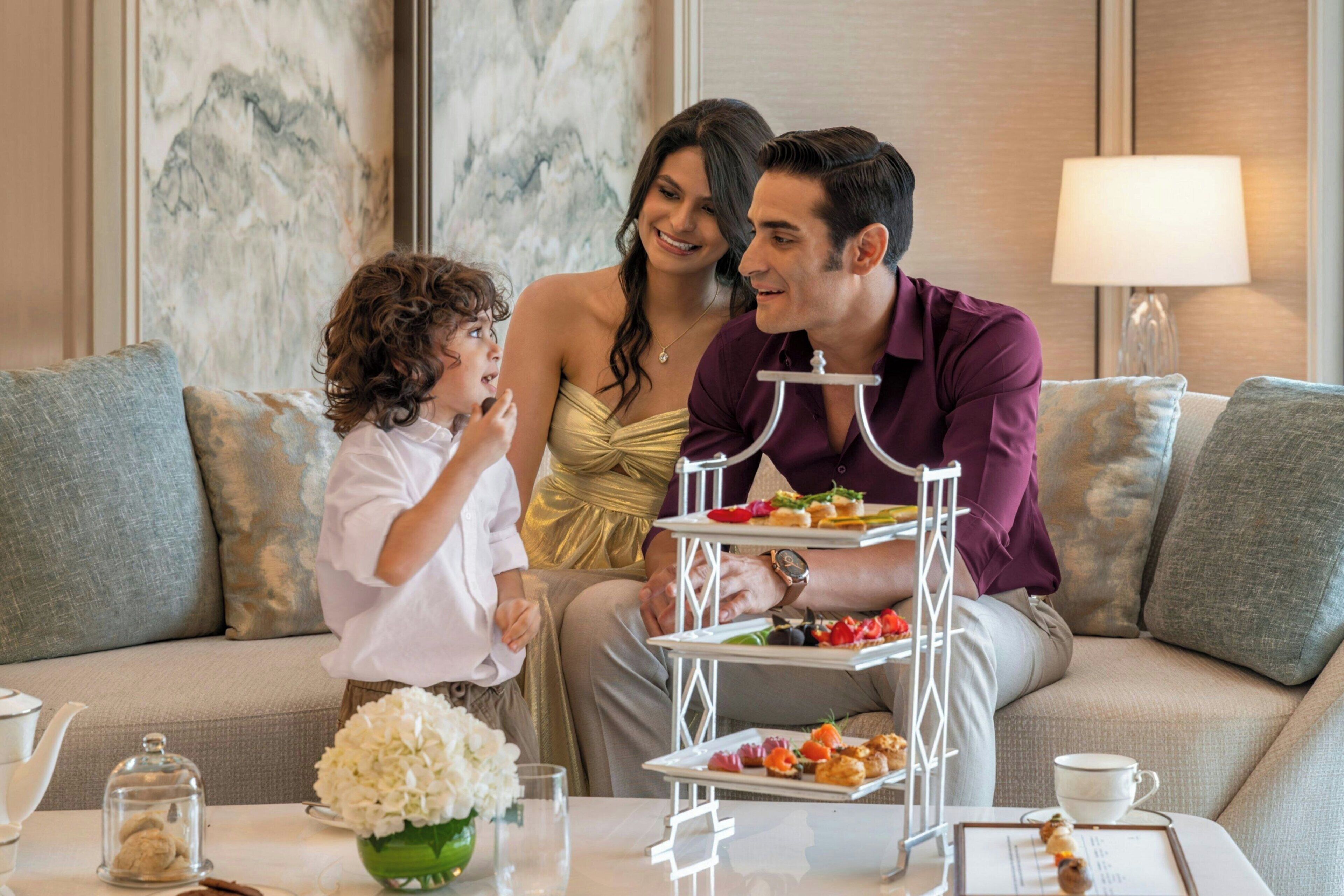 Uma família sentada em um sofá, com um suporte escalonado para sobremesas e uma mesa com um vaso de flores.
