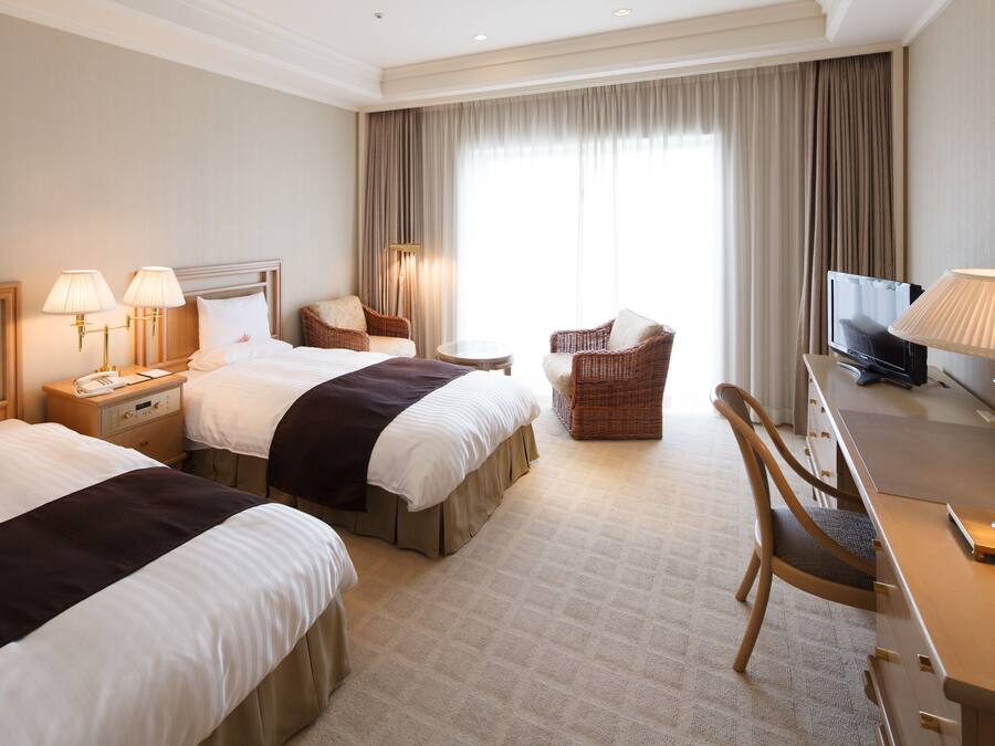 Quarto de hotel com duas camas, uma escrivaninha, uma cadeira, uma televisão e uma janela com cortinas.