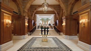 Um amplo corredor com piso de mármore, painéis de madeira ricamente decorados e um lustre central.