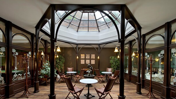 Um quarto espaçoso com pé direito alto e claraboia, com piso de madeira, várias mesas e cadeiras, e elementos decorativos como plantas e obras de arte.