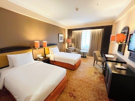 Quarto de hotel com duas camas, uma escrivaninha, uma cadeira, televisão e janela com cortinas.