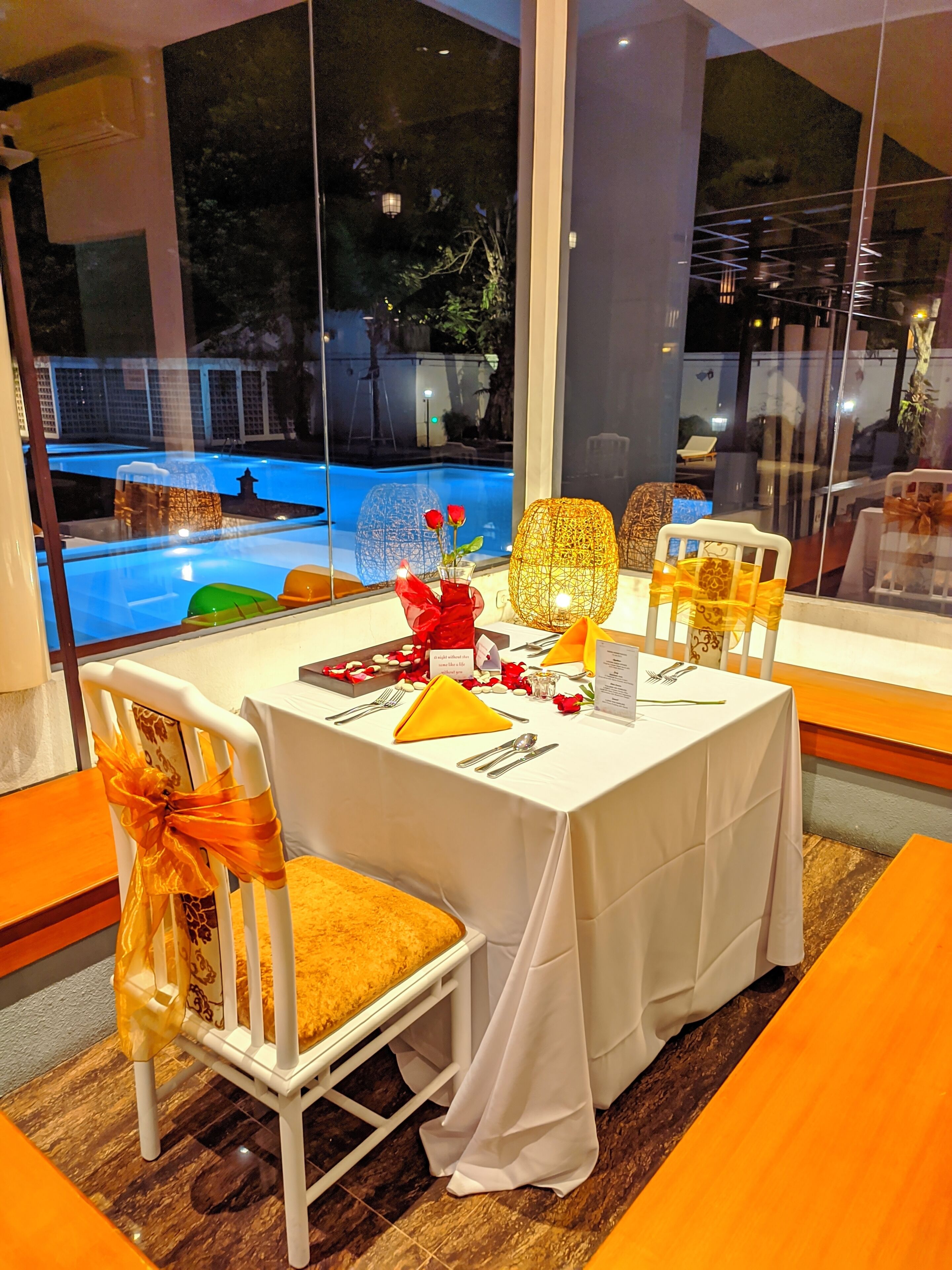 Mesa de jantar posta para dois, com toalha de mesa branca, vaso de vidro com rosas vermelhas e um lanterna decorativa.