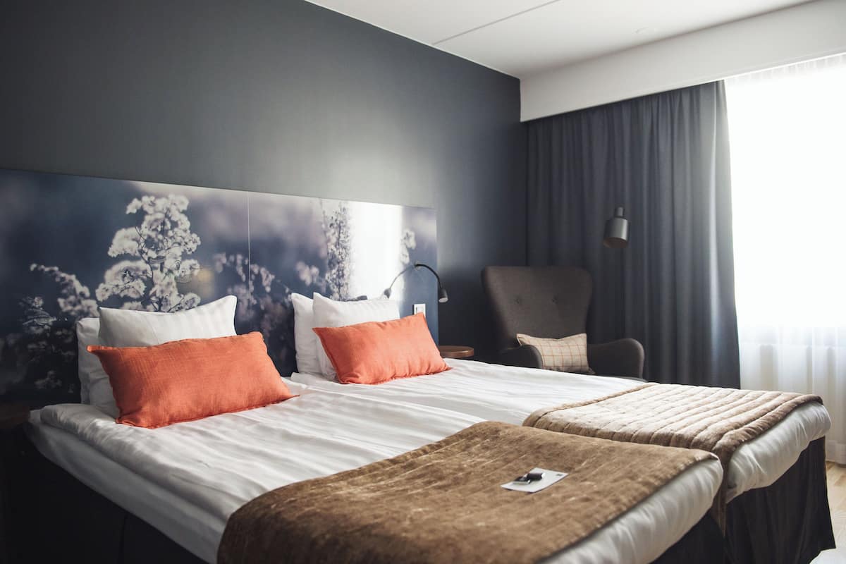 Quarto de hotel com uma cama grande, duas almofadas, mesa de cabeceira, cadeira e vista para o exterior.