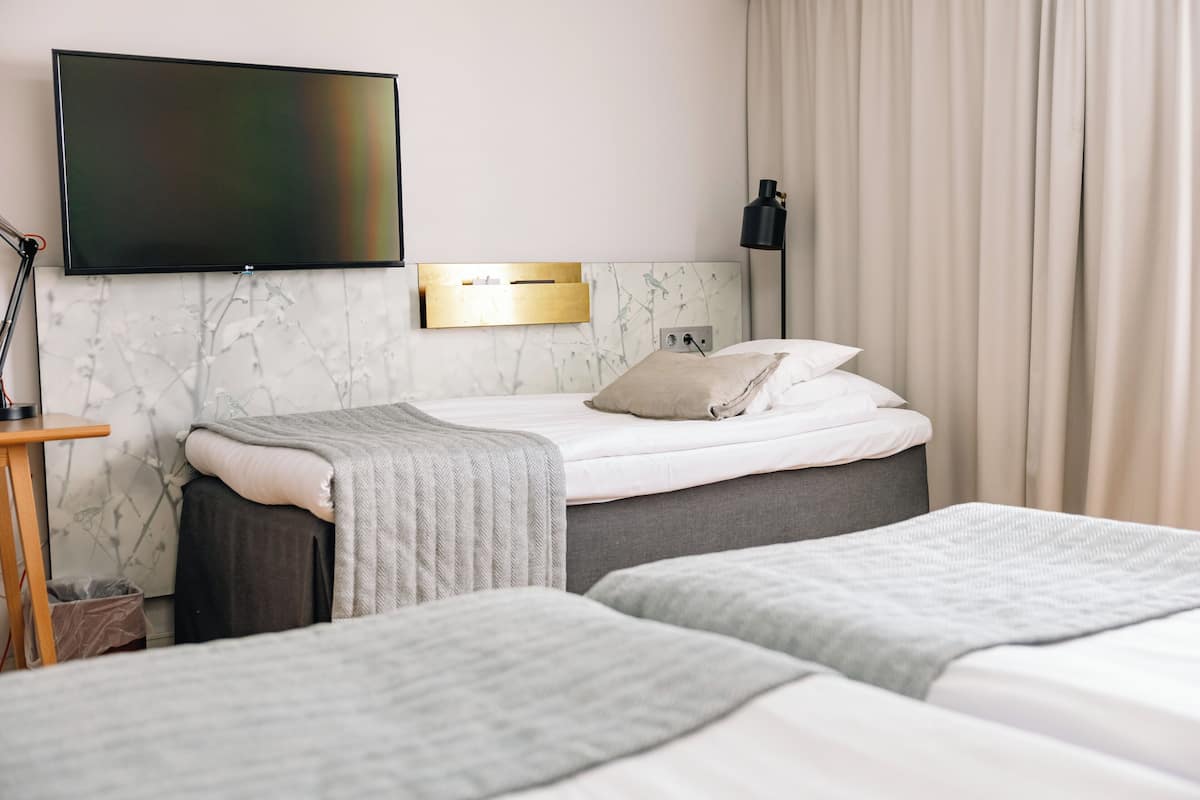 Quarto de hotel com uma televisão de tela plana grande fixada na parede, uma cama com cabeceira cinza e uma mesa de cabeceira com abajur.