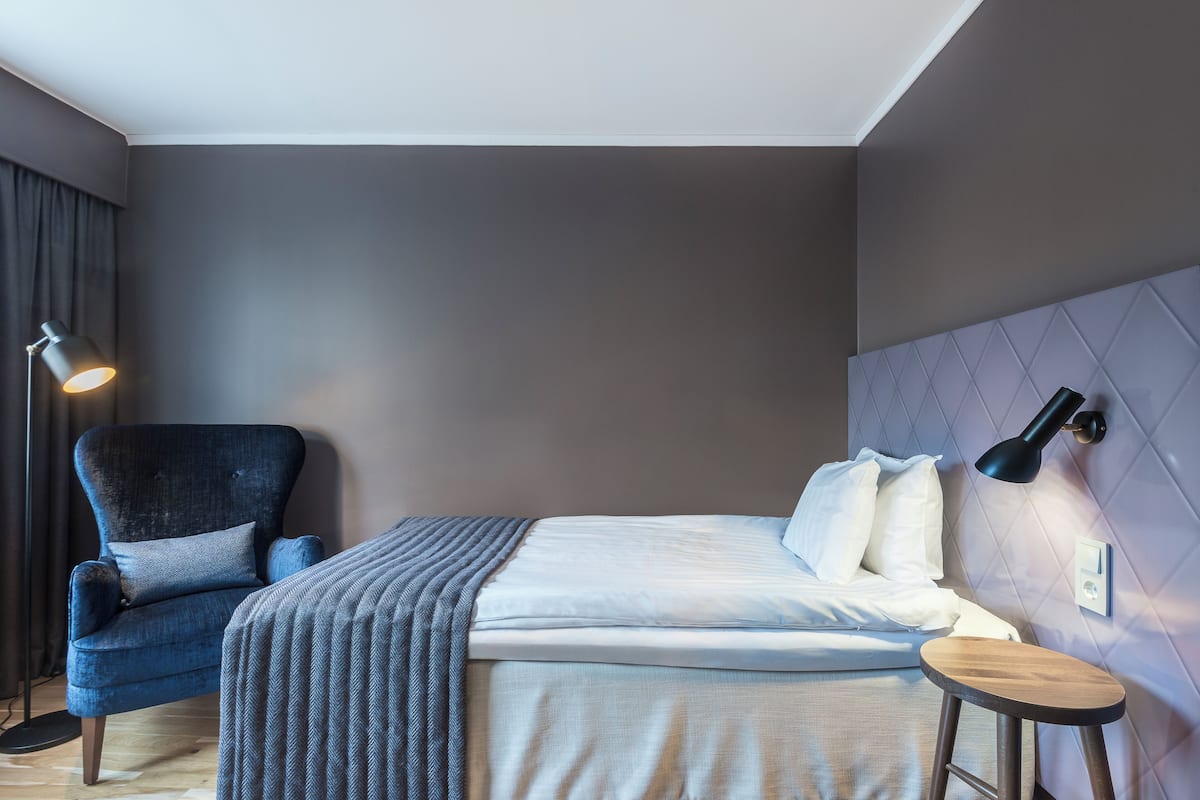 Quarto de hotel moderno com cama, uma cadeira, um banquinho e luminária fixada na parede.