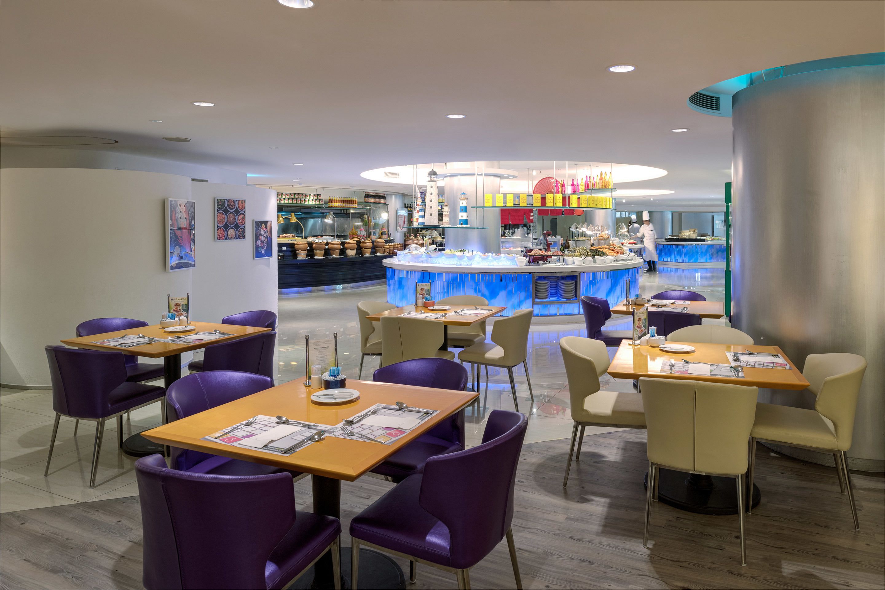 Restaurante moderno com cadeiras roxas e bege, um balcão central e diversas opções de comida expostas.