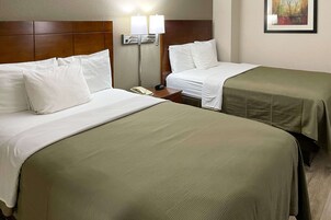Quarto de hotel com duas camas, cada uma com roupa de cama branca e colchas verdes. Há uma mesa de cabeceira com telefone e abajur entre as camas.