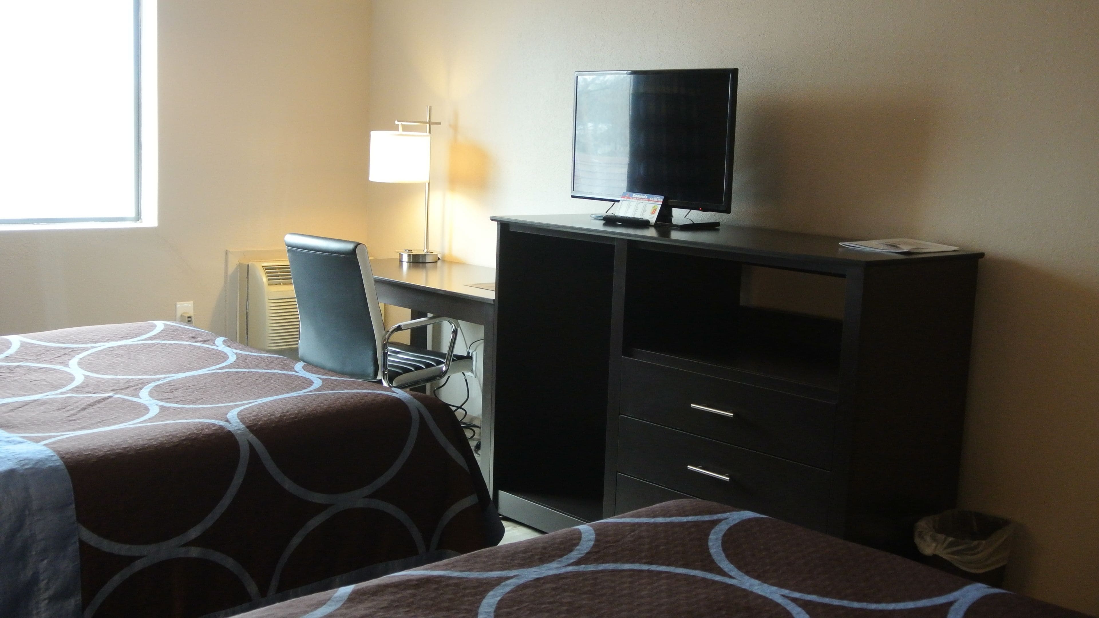 Quarto de hotel com duas camas, uma escrivaninha com computador e televisão, uma luminária e uma janela.