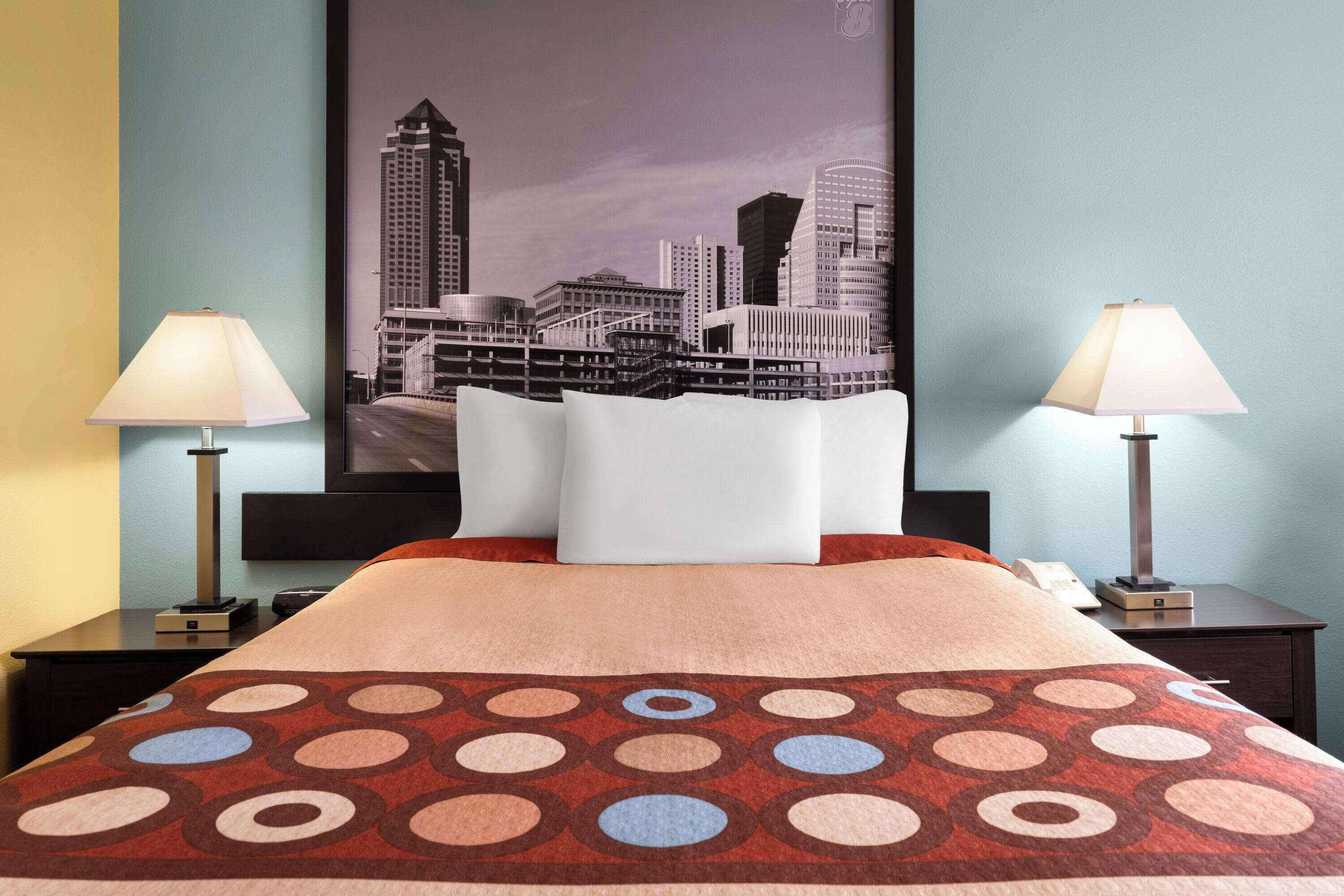 Quarto de hotel com cama, duas mesas de cabeceira, um cartaz com vista urbana na parede e um edredom estampado.