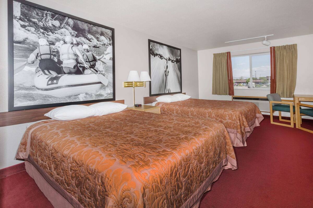 Quarto de hotel com duas camas, uma escrivaninha, uma cadeira, janela com cortinas e dois quadros na parede.