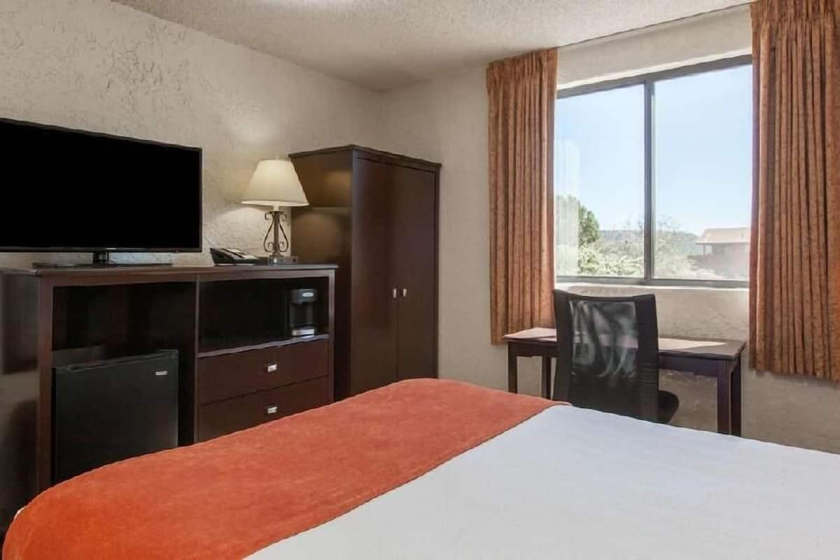 Quarto de hotel com cama, escrivaninha, televisão, abajur, guarda-roupa e janela com cortinas.