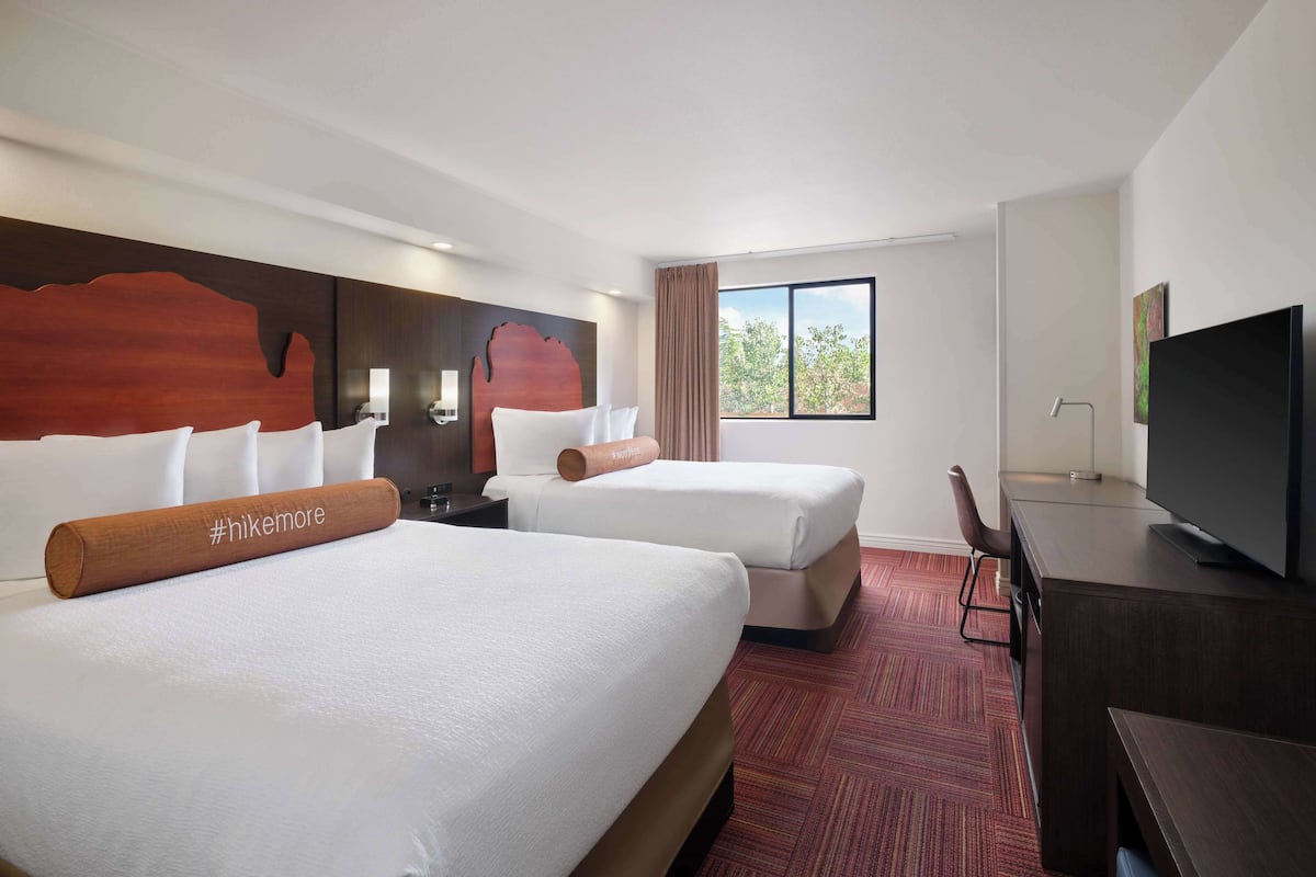 Quarto de hotel com duas camas, televisão, escrivaninha e janela com vista para árvores.