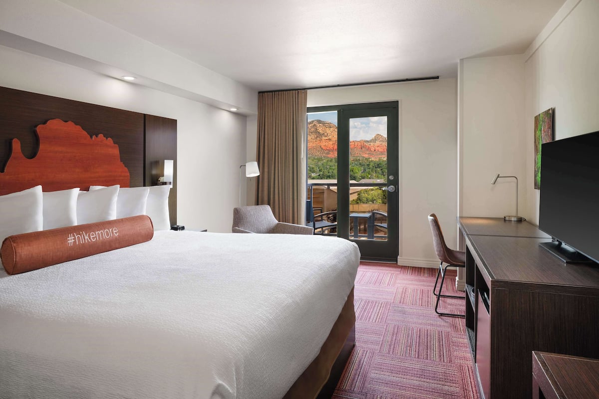 Quarto de hotel com uma cama grande, uma mesa, uma TV e uma varanda com vista para formações rochosas avermelhadas.