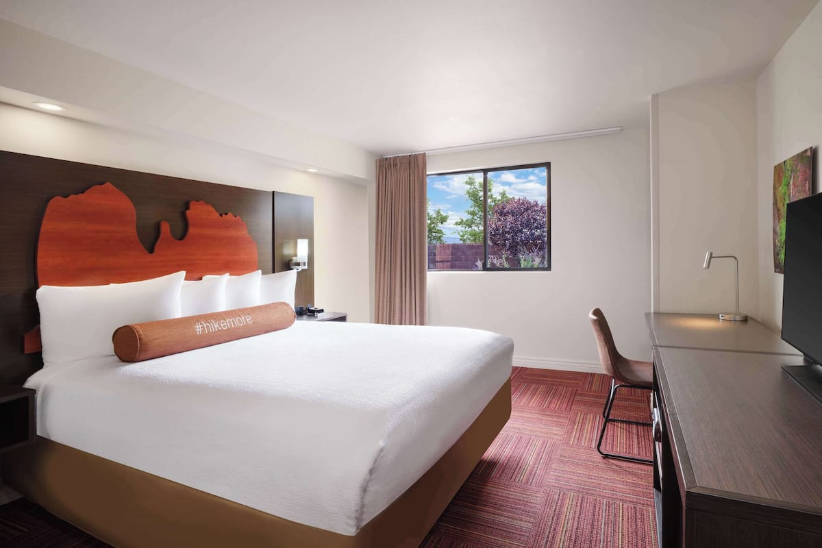 Quarto de hotel com uma cama grande, uma escrivaninha, uma cadeira, uma televisão e uma janela com vista para árvores.