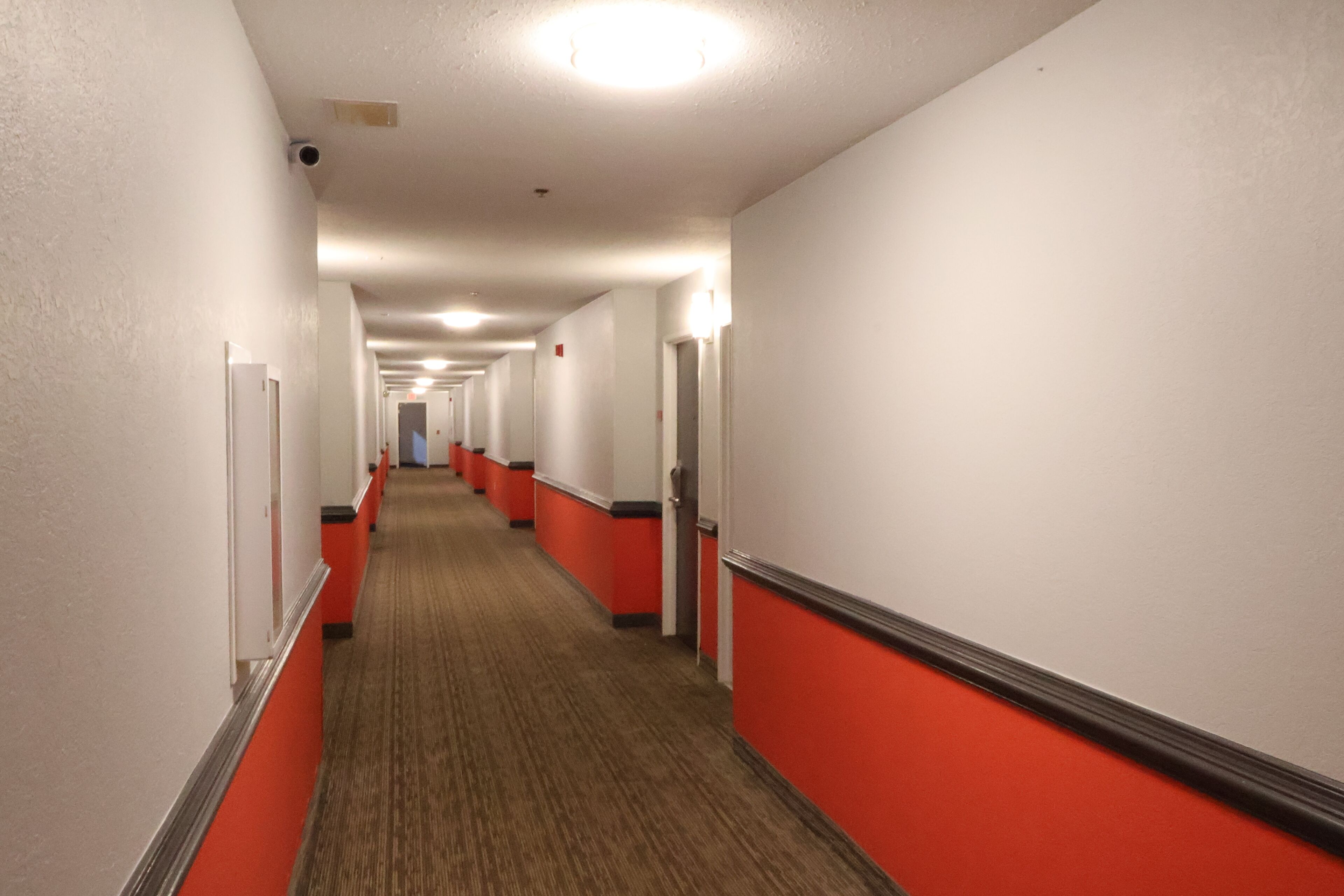 Um corredor comprido e estreito, com paredes vermelhas e brancas, pisos cobertos de carpete e portas em ambos os lados.