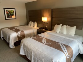 Quarto de hotel com duas camas, uma mesa de cabeceira com abajur e um quadro na parede.