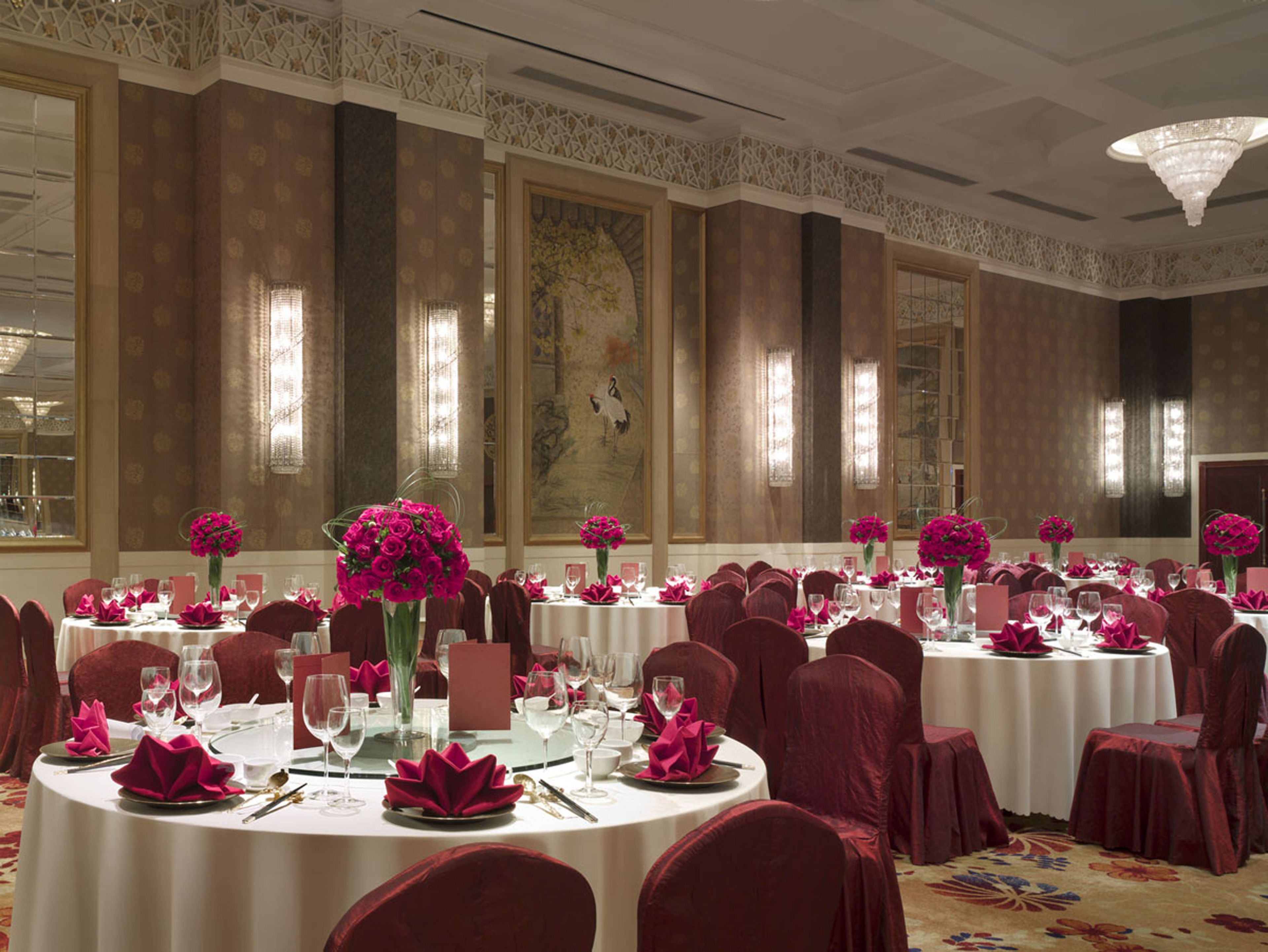 Uma sala de banquetes com mesas redondas preparadas para refeições, decorada com flores cor-de-rosa e guardanapos vermelhos.