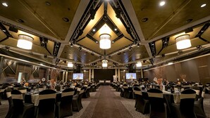 Uma grande sala de conferências com fileiras de mesas e cadeiras, equipada com iluminação pendente e um palco na frente.