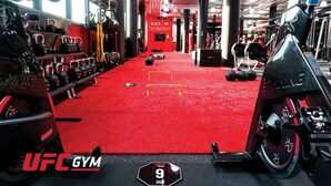 Uma academia com piso vermelho, equipamentos de musculação e uma placa indicando ser a UFC Gym 9.