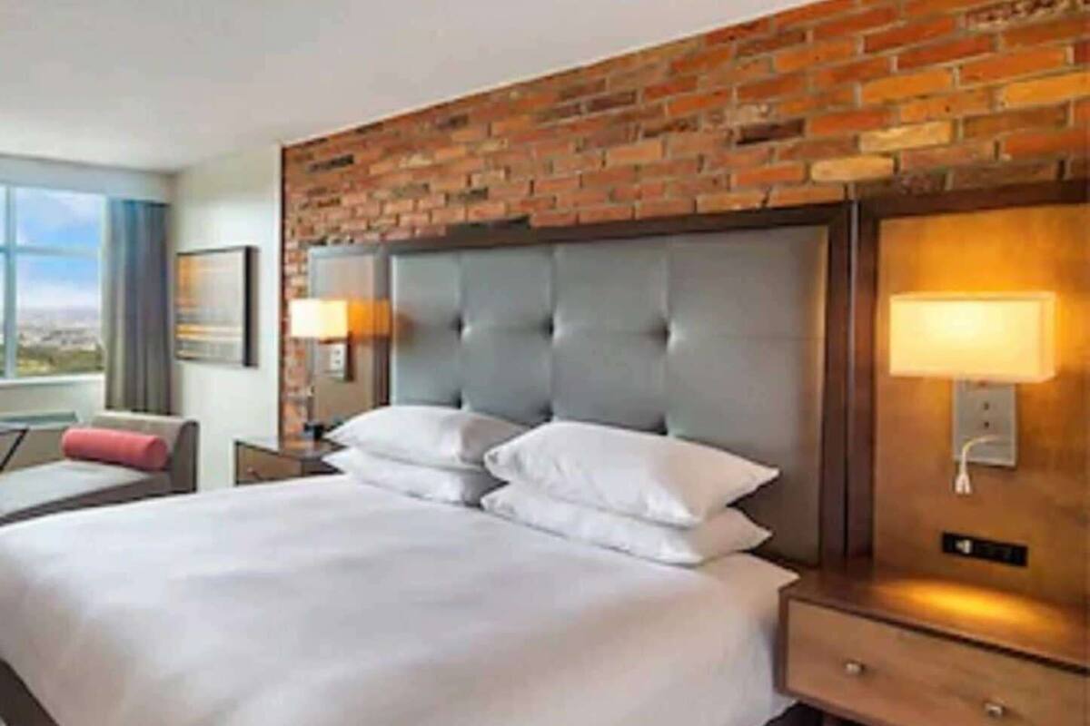 Quarto de hotel com uma cama grande, parede de tijolos, mesa de cabeceira com abajur e vista pela janela.