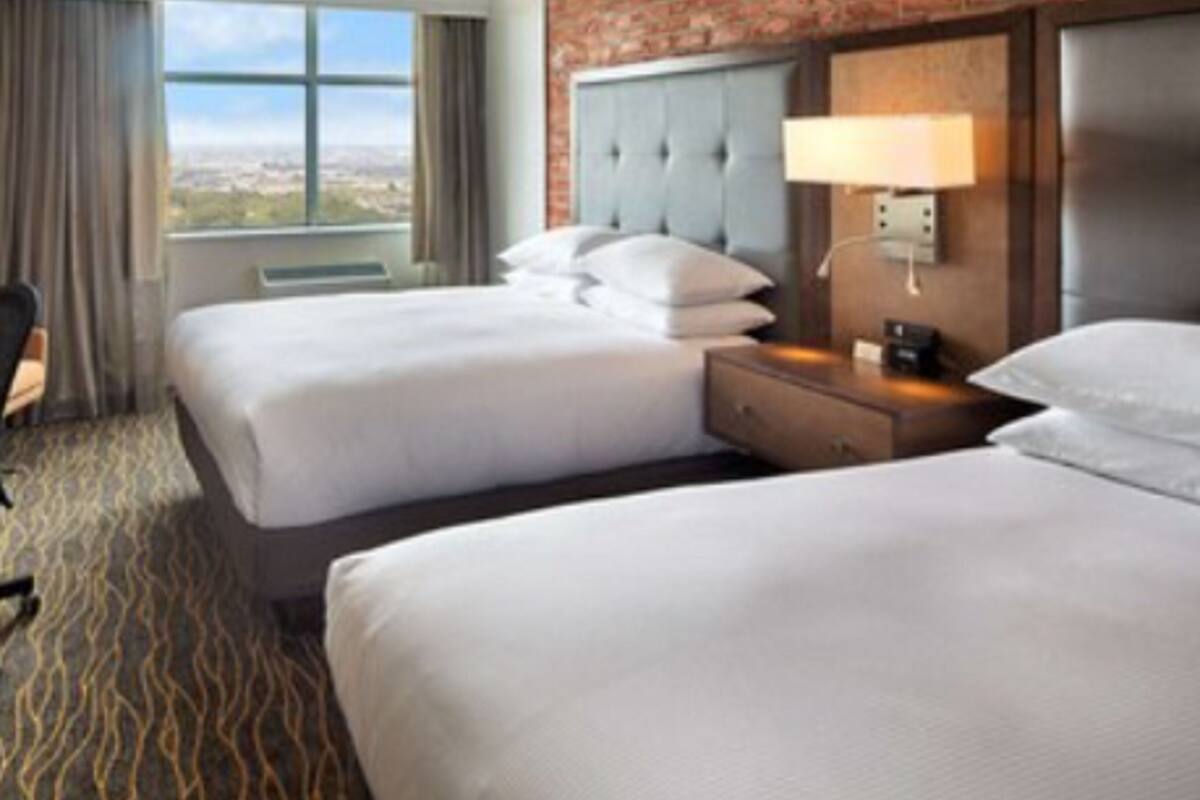 Quarto de hotel com duas camas, parede de tijolos decorativa, grande janela com vista para a cidade e escrivaninha com luminária.