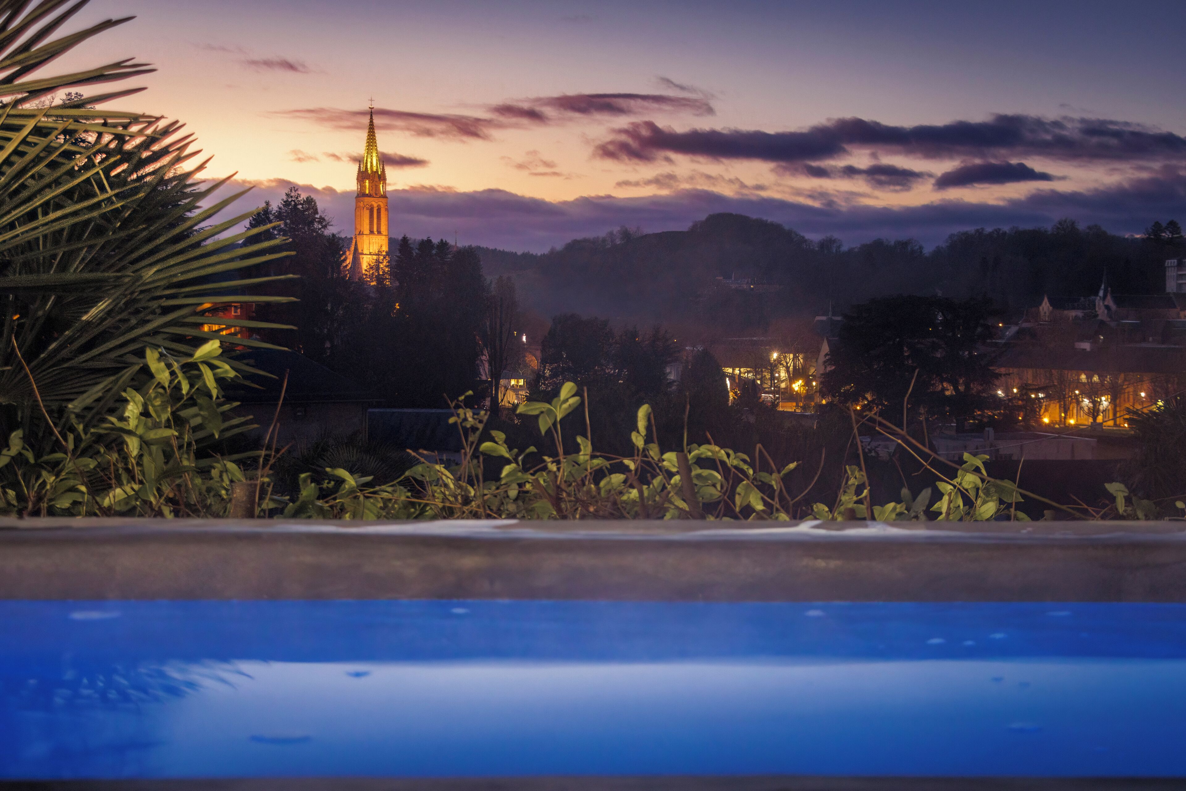 Vista de um campanário de igreja ao entardecer, com edifícios e vegetação ao redor.