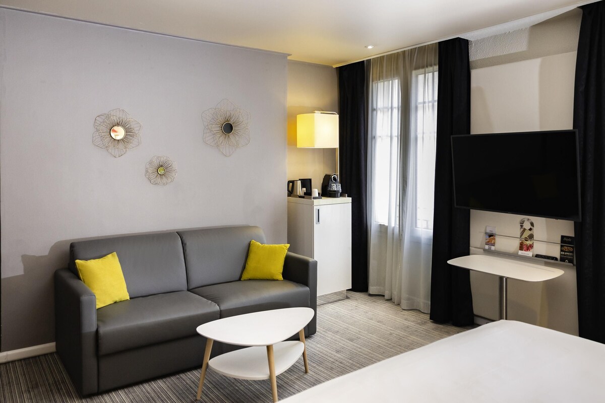 Quarto de hotel moderno com sofá cinza, uma mesinha branca, uma cama, TV e minibar.