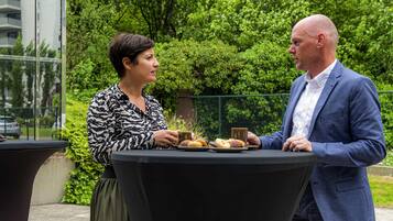 Duas pessoas num espaço ao ar livre, numa mesa com comida e bebidas, rodeadas por vegetação e um edifício ao fundo.