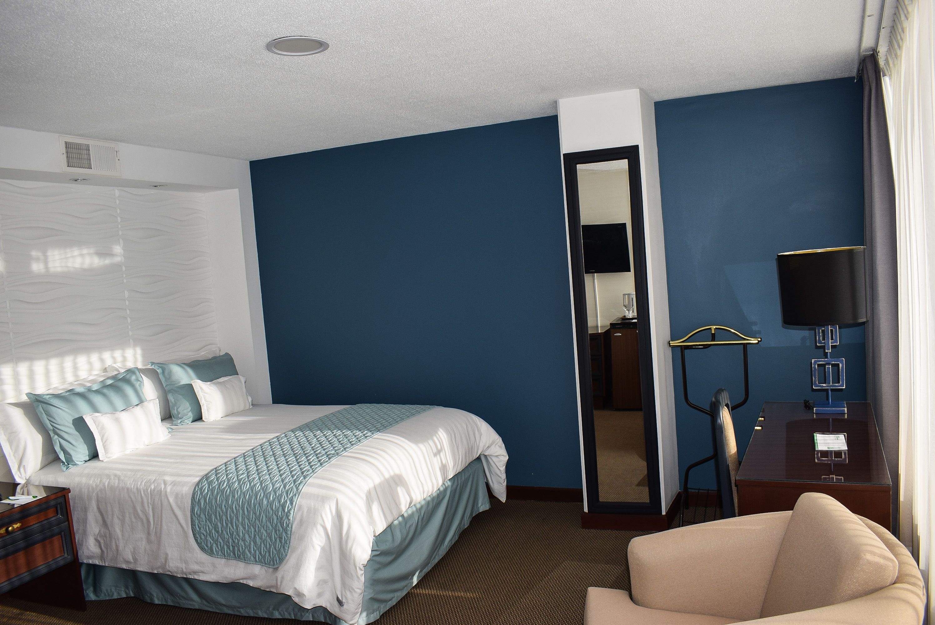 Quarto de hotel com cama, poltrona, escrivaninha com computador e espelho.