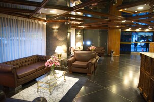 Um lobby com um sofá de couro marrom, uma poltrona marrom, uma mesa de centro de vidro com um vaso de flores e um balcão de recepção.