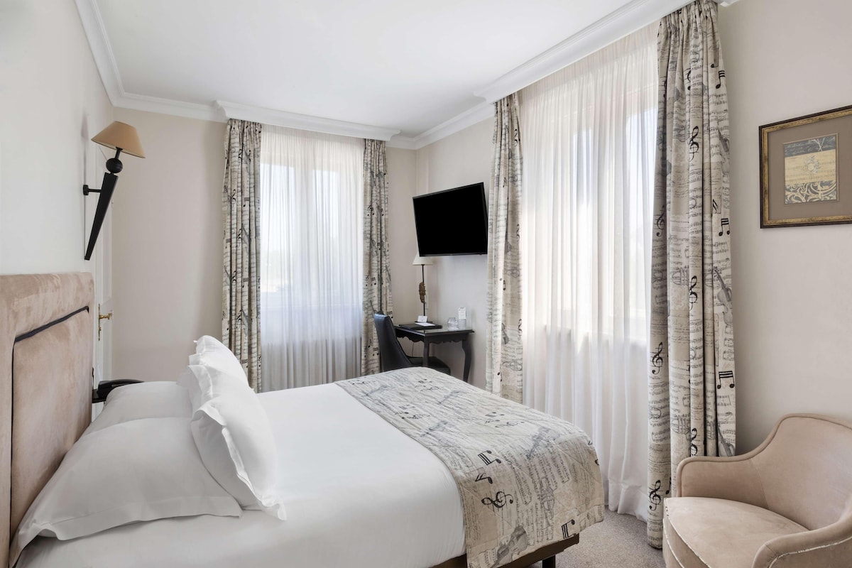 Quarto de hotel com cama, escrivaninha, televisão de tela plana, cadeira e cortinas com padrão de nota musical.
