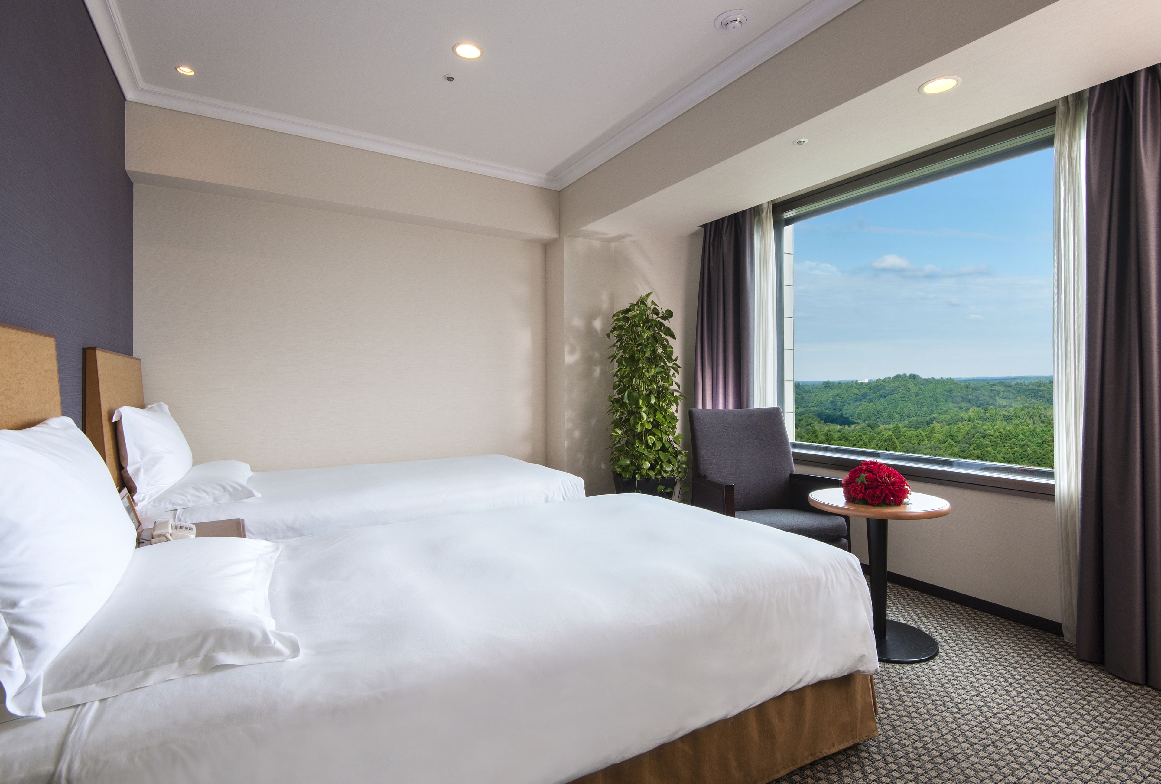 Quarto de hotel com uma cama grande, uma pequena mesa com uma flor, uma cadeira e vista para o exterior.