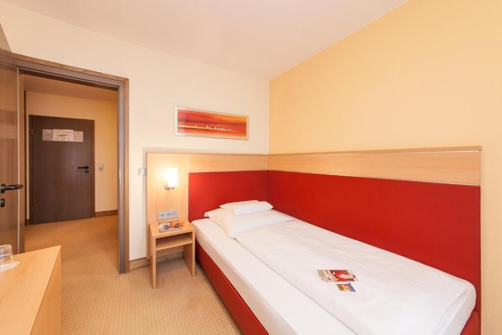 Quarto de hotel com uma cama de solteiro, uma mesa de cabeceira com abajur, uma pequena mesa com um copo de água e uma porta que dá para outro quarto.