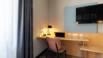 Quarto de hotel com escrivaninha, cadeira, luminária, televisão de tela plana e janela com cortinas.