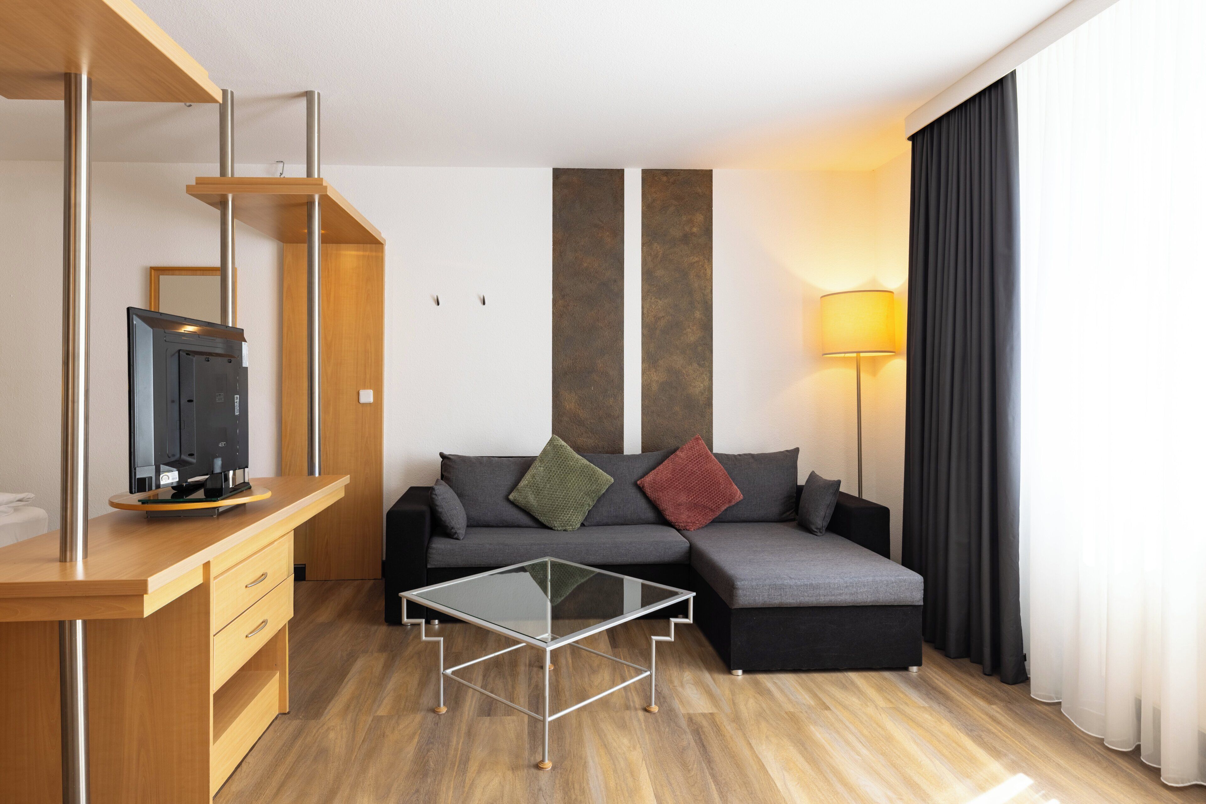 Quarto de hotel moderno com sofá, mesa de centro de vidro, escrivaninha e televisão de tela plana, além de luminária.