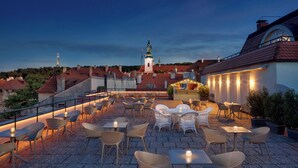 Terraço no telhado com área de estar ao ar livre e bar, com vista para uma cidade com edifícios históricos e uma torre.