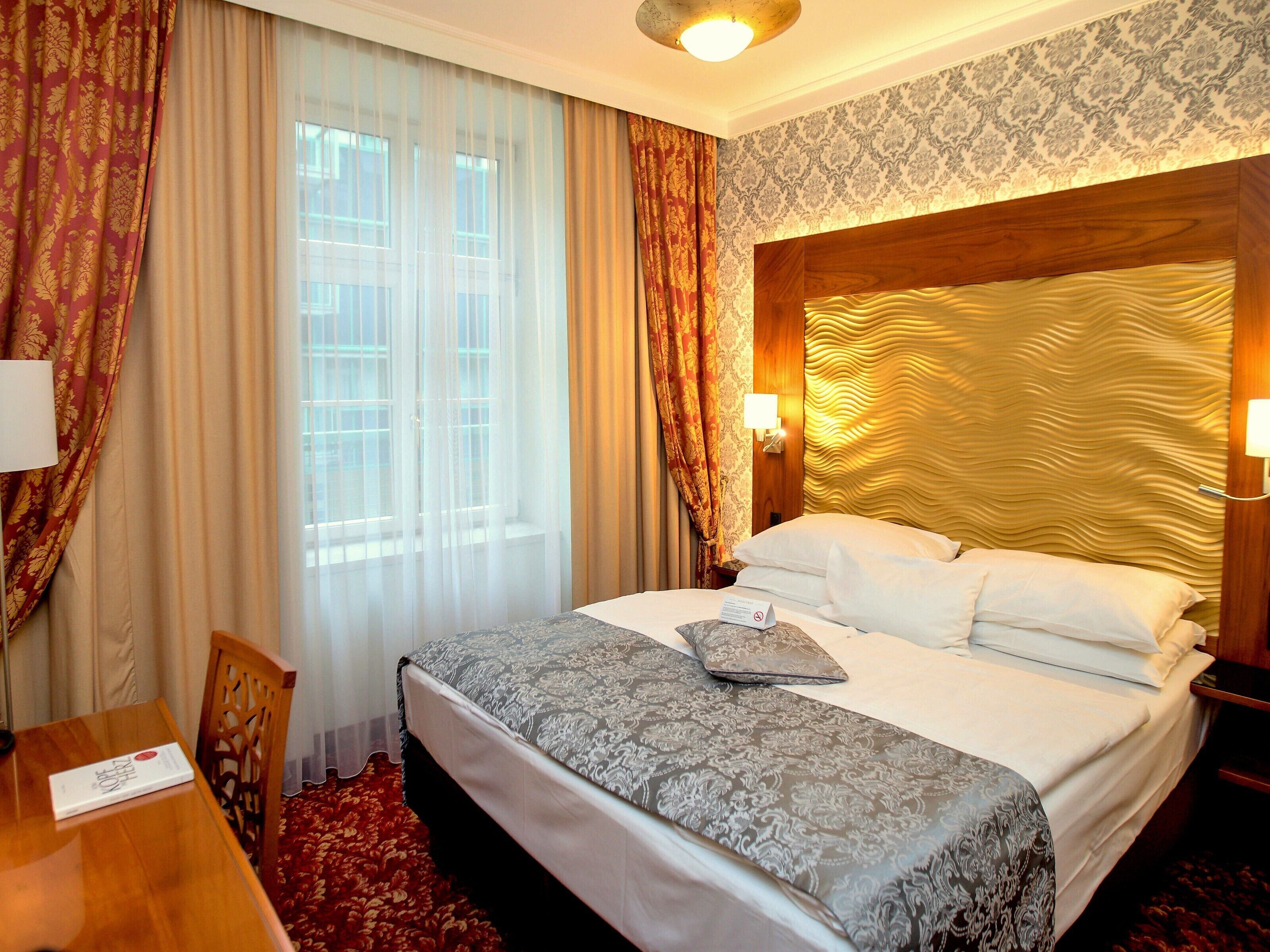 Quarto de hotel com cama, escrivaninha e cadeira. Há uma janela com cortinas, uma parede com papel de parede estampado e uma cabeceira com design exclusivo.