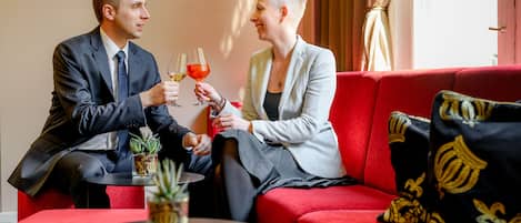 Um homem e uma mulher sentados num sofá vermelho, segurando copos de vinho. Há uma pequena mesa com uma planta em vaso à frente deles.