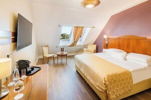 Quarto de hotel com cama, escrivaninha com luminária, cadeira, televisão e janela com cortinas.