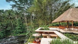 Uma villa moderna com piscina, área de estar externa e estrutura de telhado de palha, cercada por vegetação exuberante e um rio.