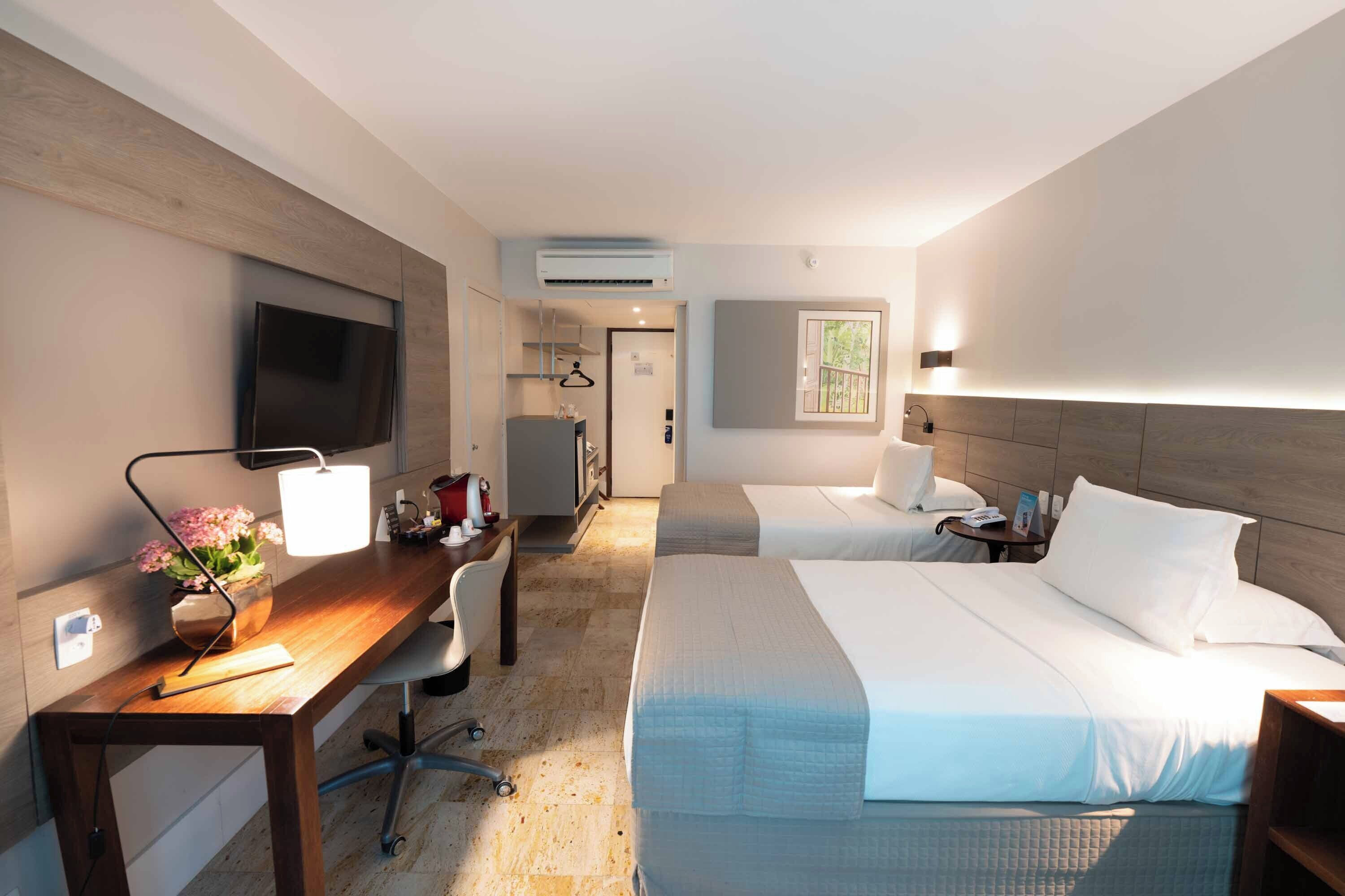 Quarto de hotel com escrivaninha de madeira, duas camas, televisão, luminária e uma pequena cozinha.