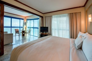 Quarto de hotel com uma cama grande, televisão, escrivaninha com cadeira e vista para o mar através de grandes janelas.