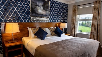 Quarto de hotel com uma cama grande, duas mesas de cabeceira com luminárias, um encosto de cama, parede estampada, um quadro e uma janela com cortinas.