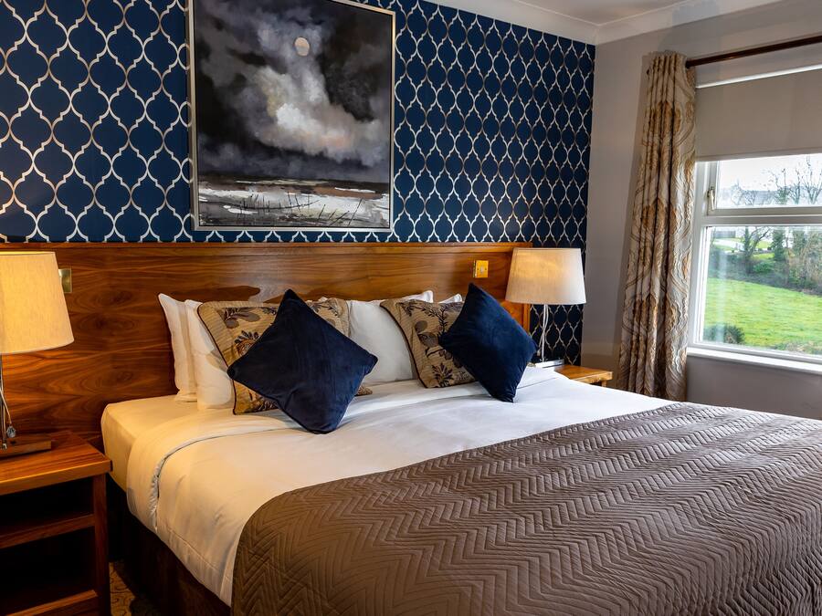 Quarto de hotel com uma cama grande, duas mesas de cabeceira com luminárias, um encosto de cama, parede estampada, um quadro e uma janela com cortinas.