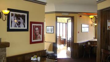 Um restaurante com área de refeições, mesas e cadeiras de madeira, quadros decorando as paredes e vista para o interior do salão através de uma porta aberta.