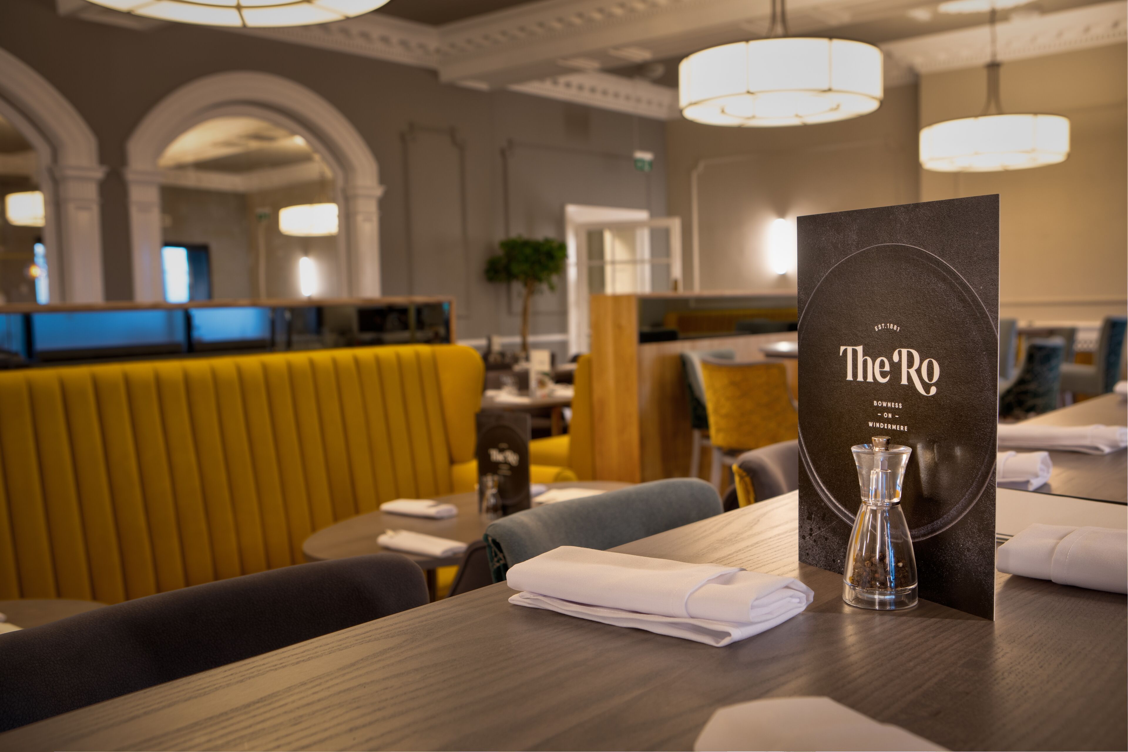Interior de restaurante com mesa posta para dois, um suporte para cardápio e área de assentos em um banco amarelo.